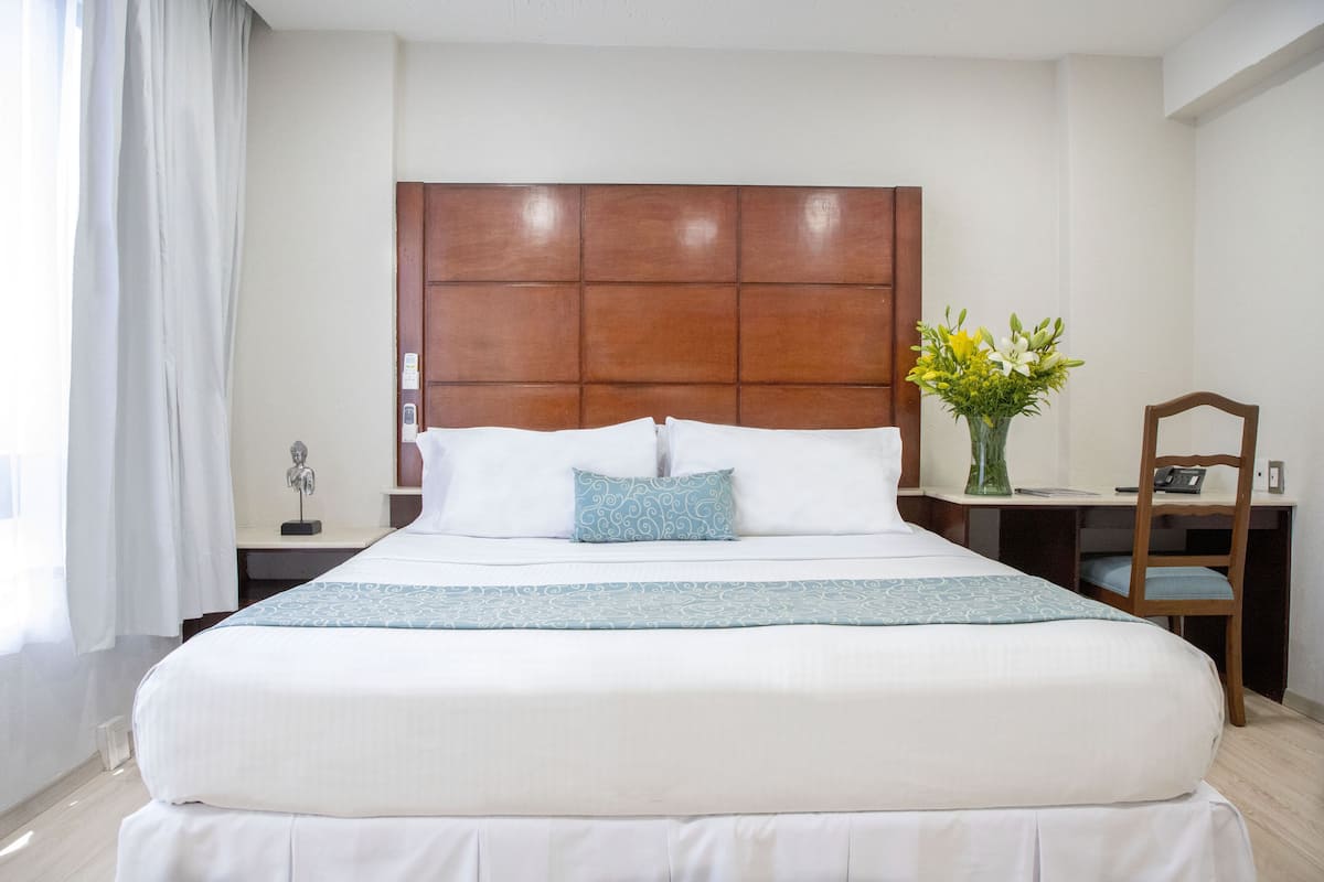Quarto de hotel com uma cama grande, cabeceira de madeira, escrivaninha com cadeira e um vaso com flores.