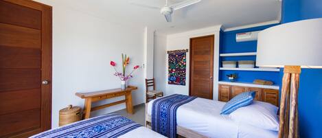 Quarto com duas camas, um ventilador de teto, uma escrivaninha, uma cadeira, uma mesa pequena com um vaso de flores e uma prateleira na parede com objetos decorativos.