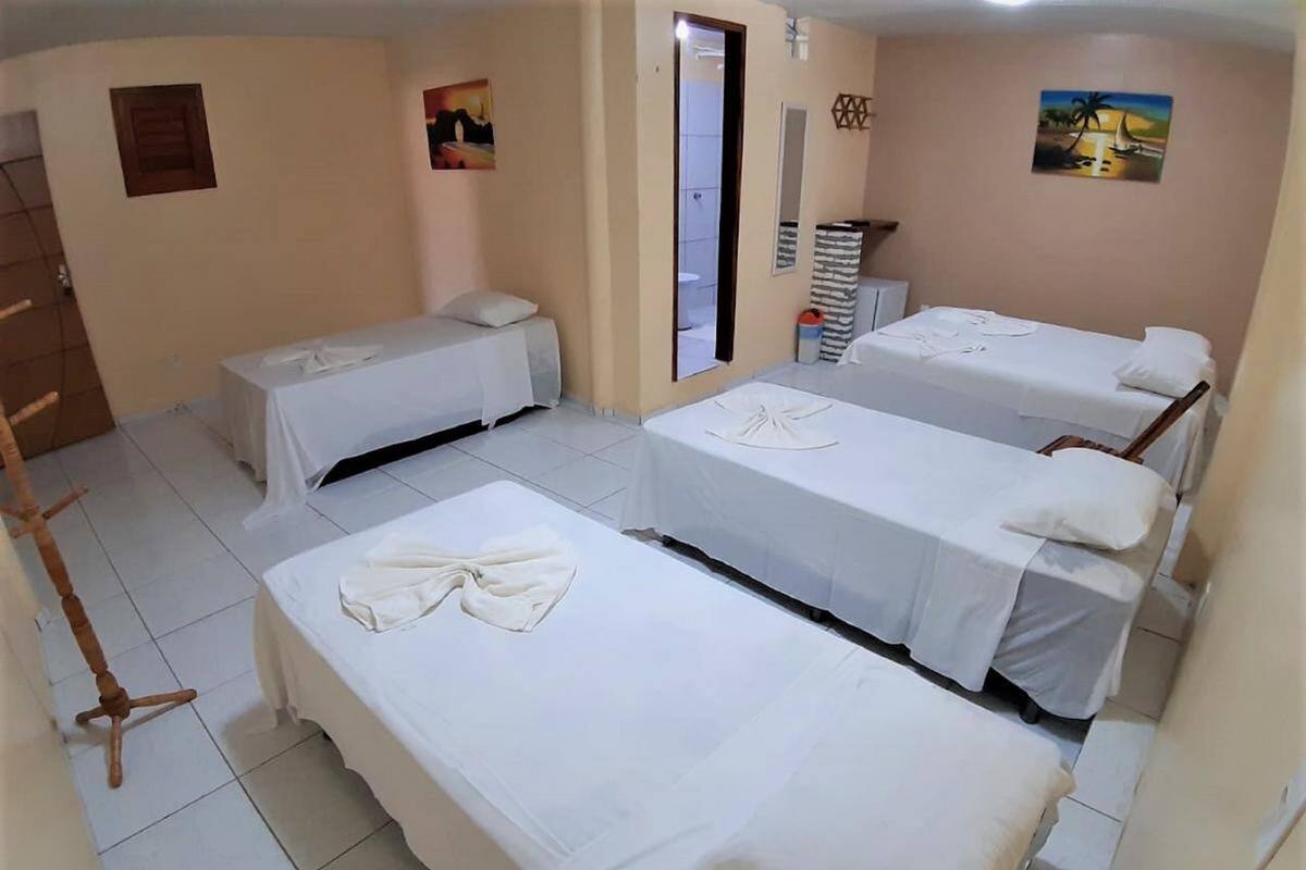 Quarto de hotel com três camas, uma cadeira de madeira e uma mesa pequena.