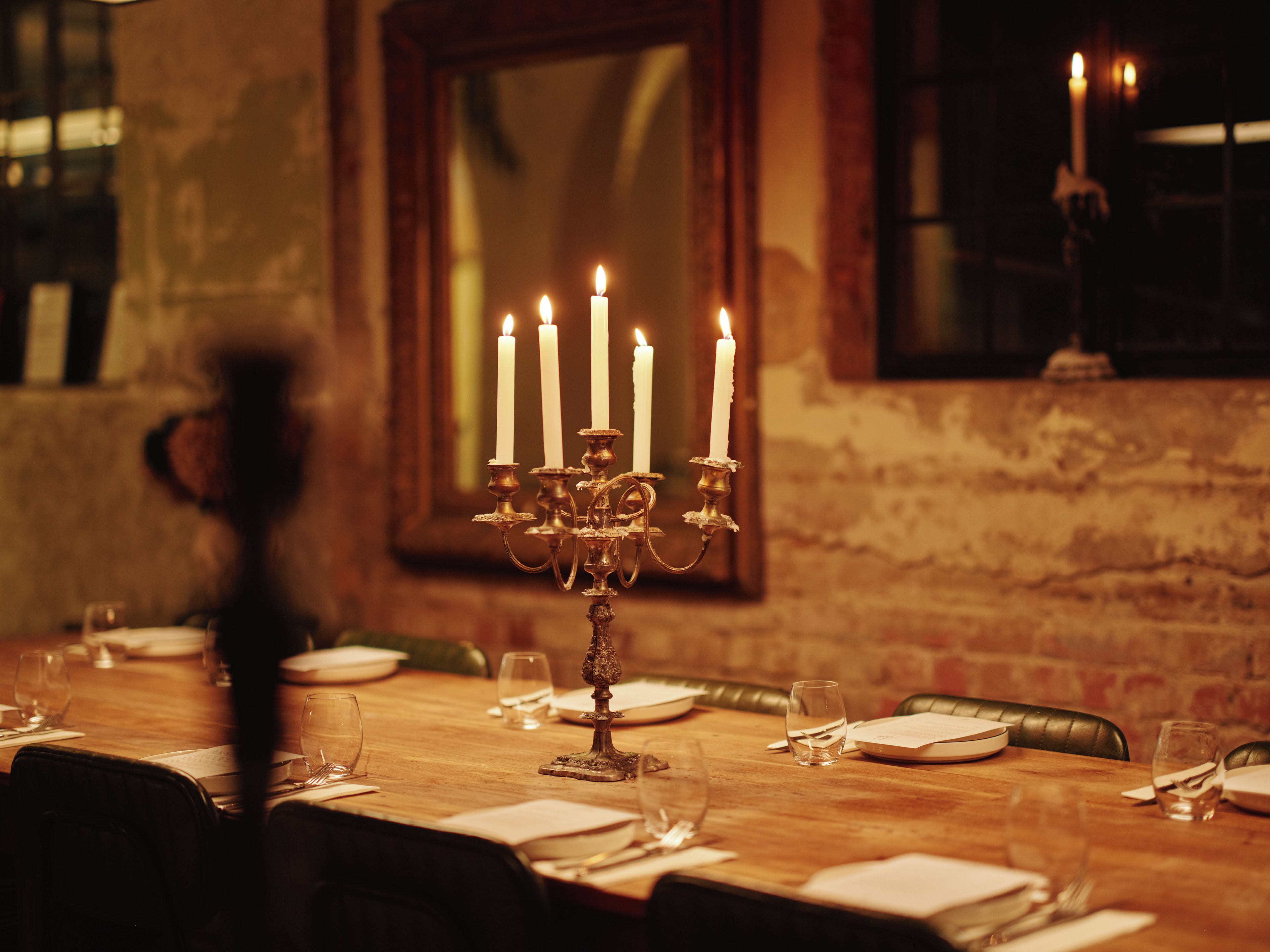 Uma mesa iluminada por velas com castiçal, copos, pratos e um cardápio.