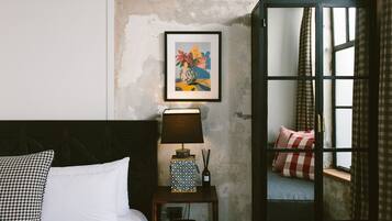 Quarto com cama, mesa de cabeceira com abajur, uma obra de arte emoldurada e janela com cortinas.