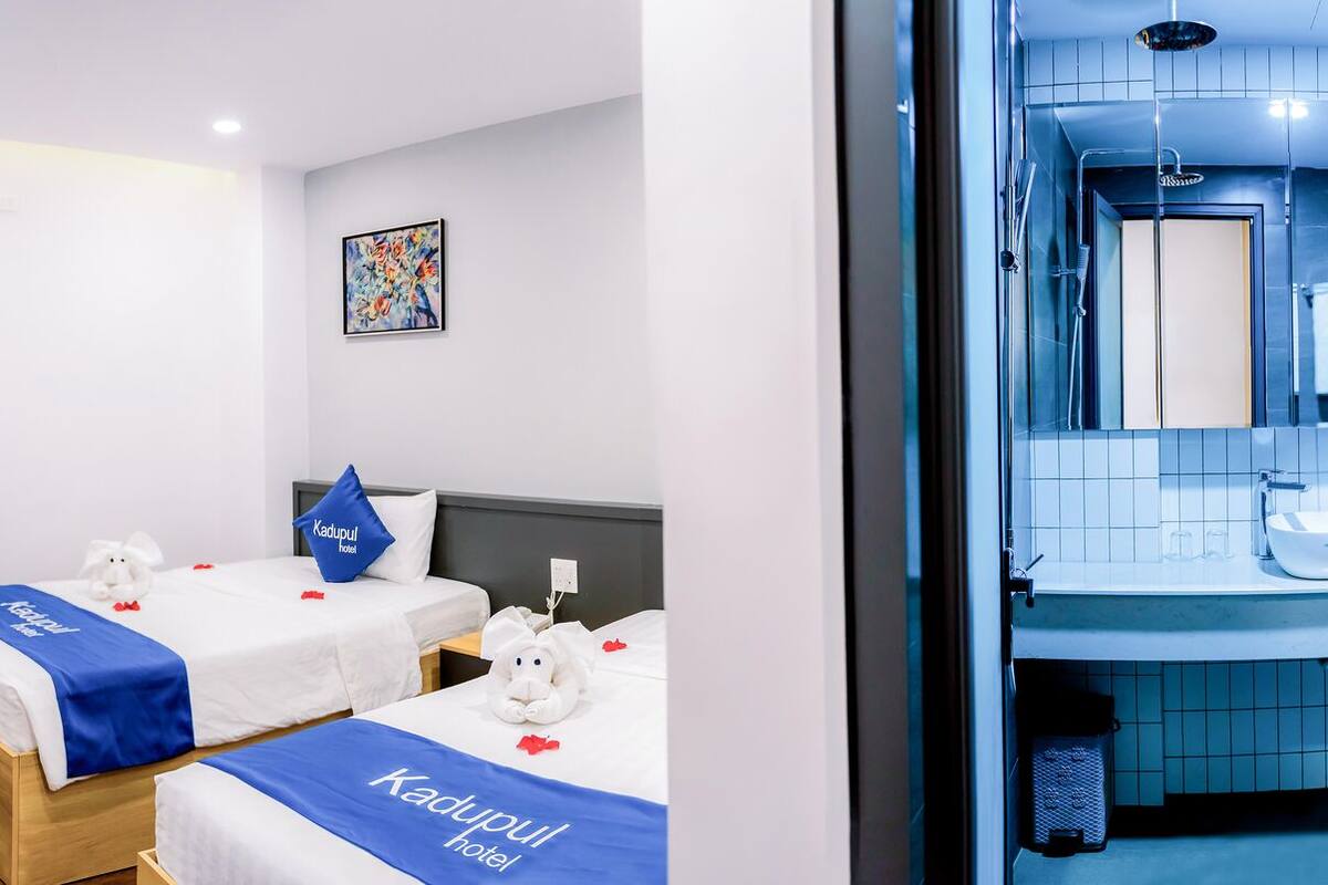 Quarto de hotel com duas camas, banheiro com chuveiro e uma pequena área de estar.