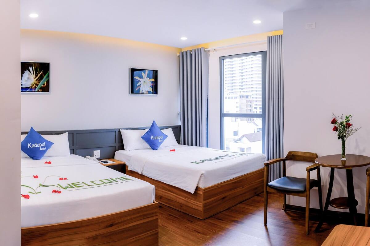 Quarto de hotel com duas camas, chuveiro, uma pequena mesa com cadeiras e uma janela com cortinas.