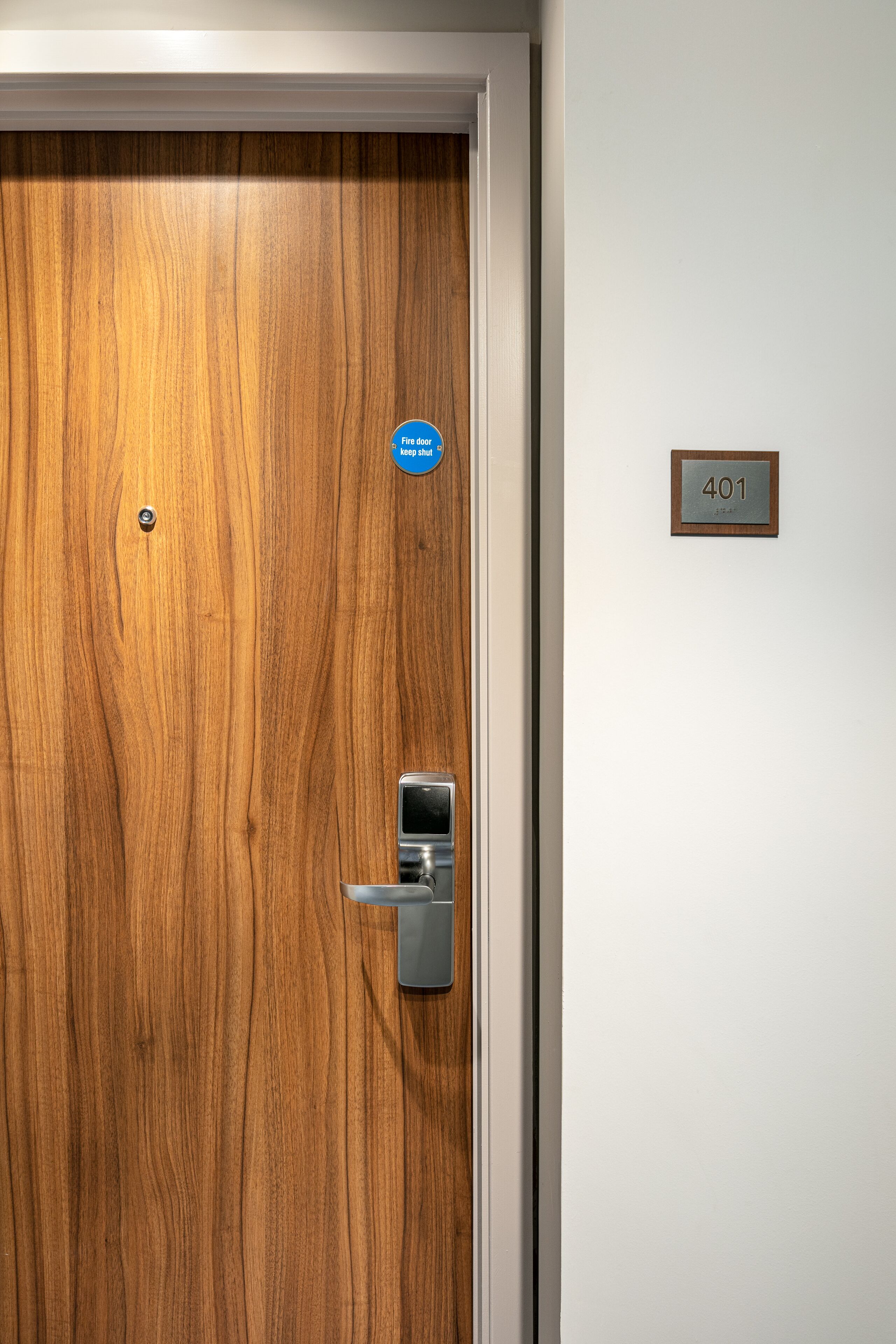 Uma porta de madeira com uma fechadura digital e uma placa azul que indica porta corta-fogo. Uma placa com o número "401" é visível na parede.
