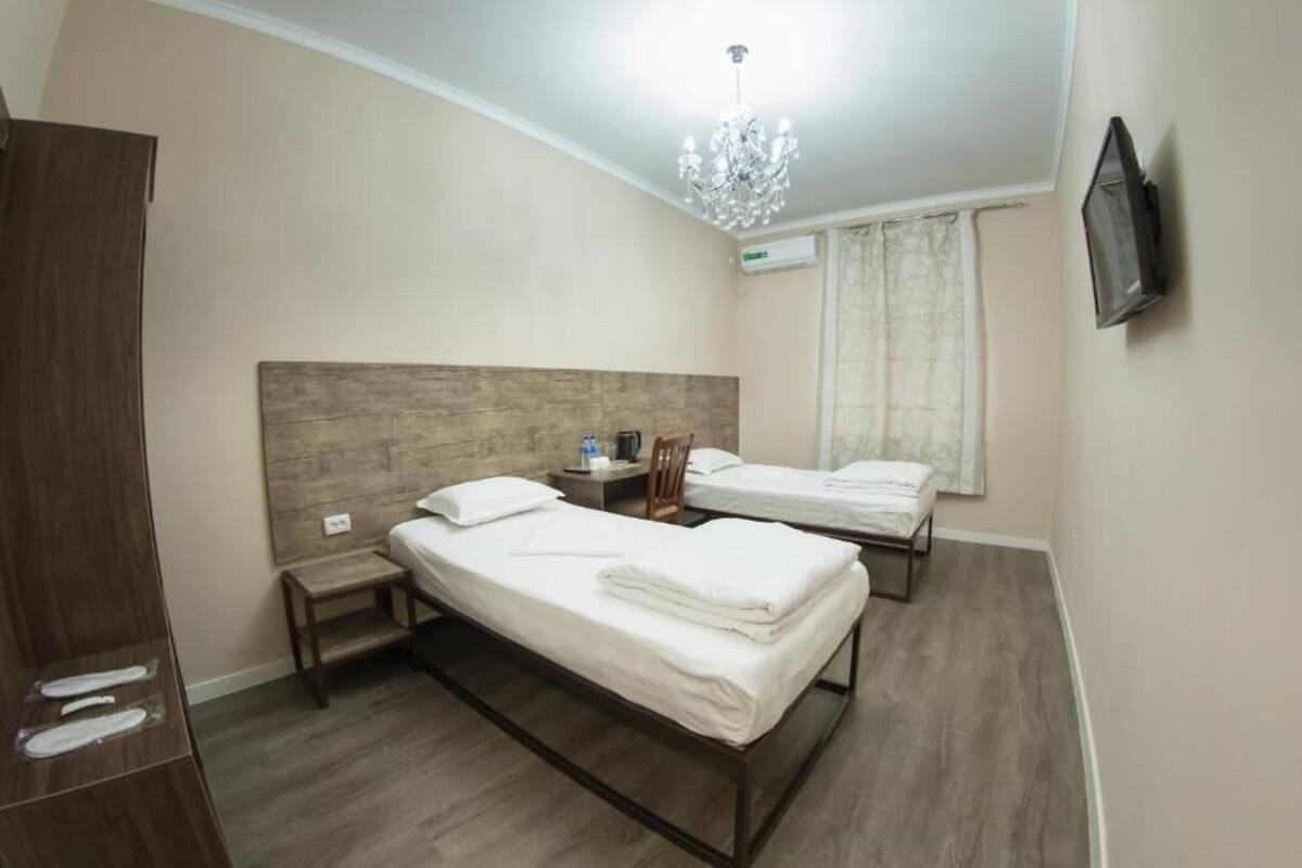 Quarto com duas camas, cabeceira de madeira, mesa de cabeceira, pequena mesa, cadeira, lustre, televisão na parede e janela com cortinas.