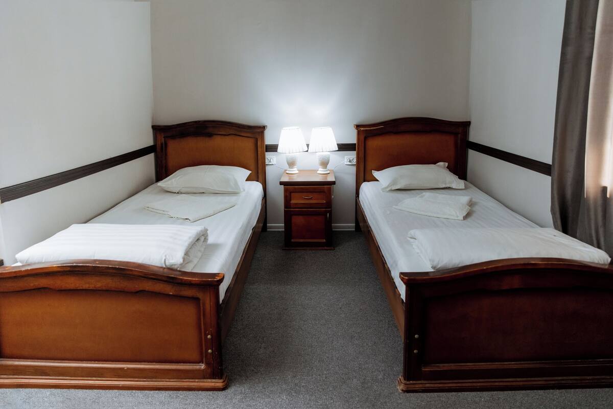 Quarto de hotel com duas camas de madeira, uma mesa de cabeceira com luminárias e piso coberto por carpete.