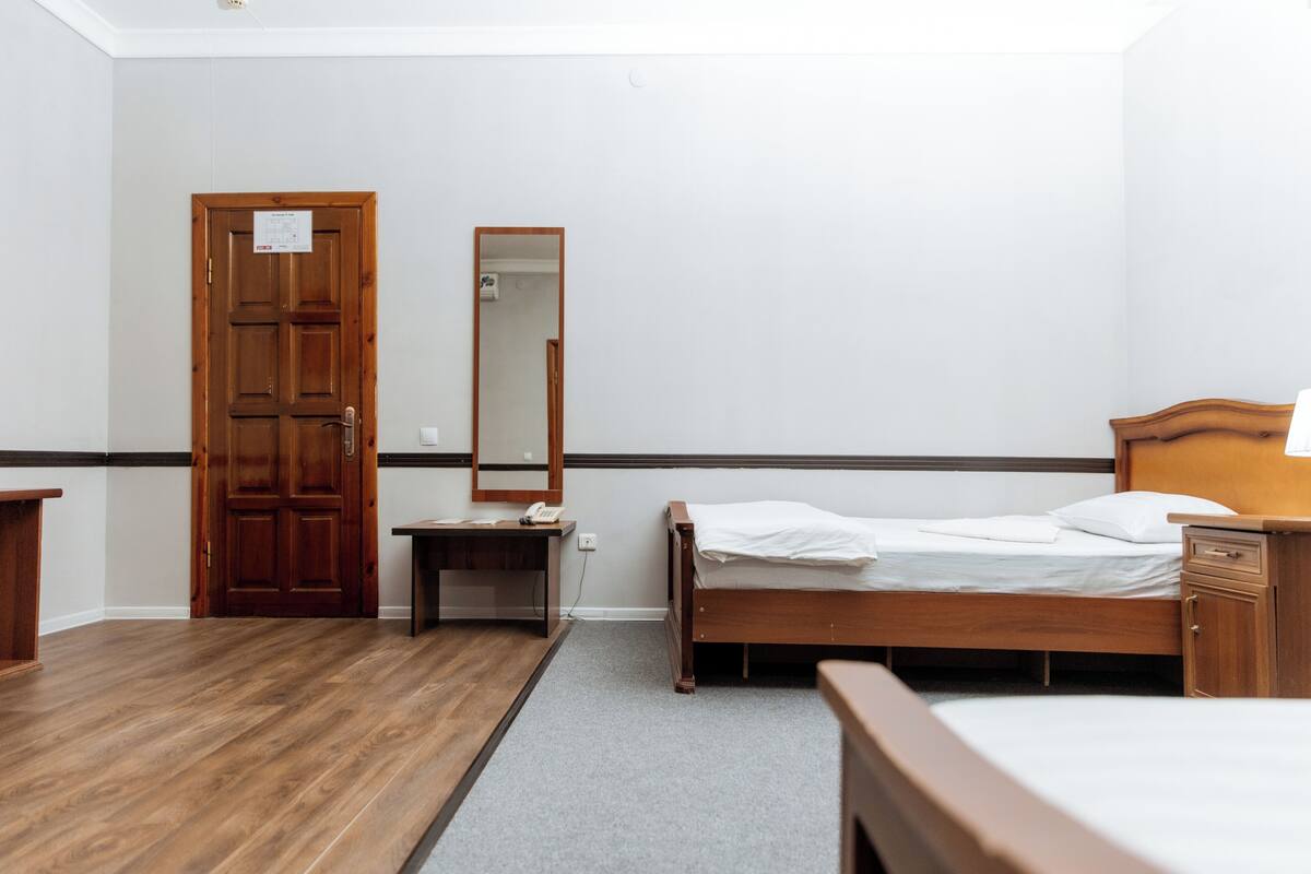 Um quarto com porta de madeira, mesa de cabeceira, cama, espelho e criado-mudo.