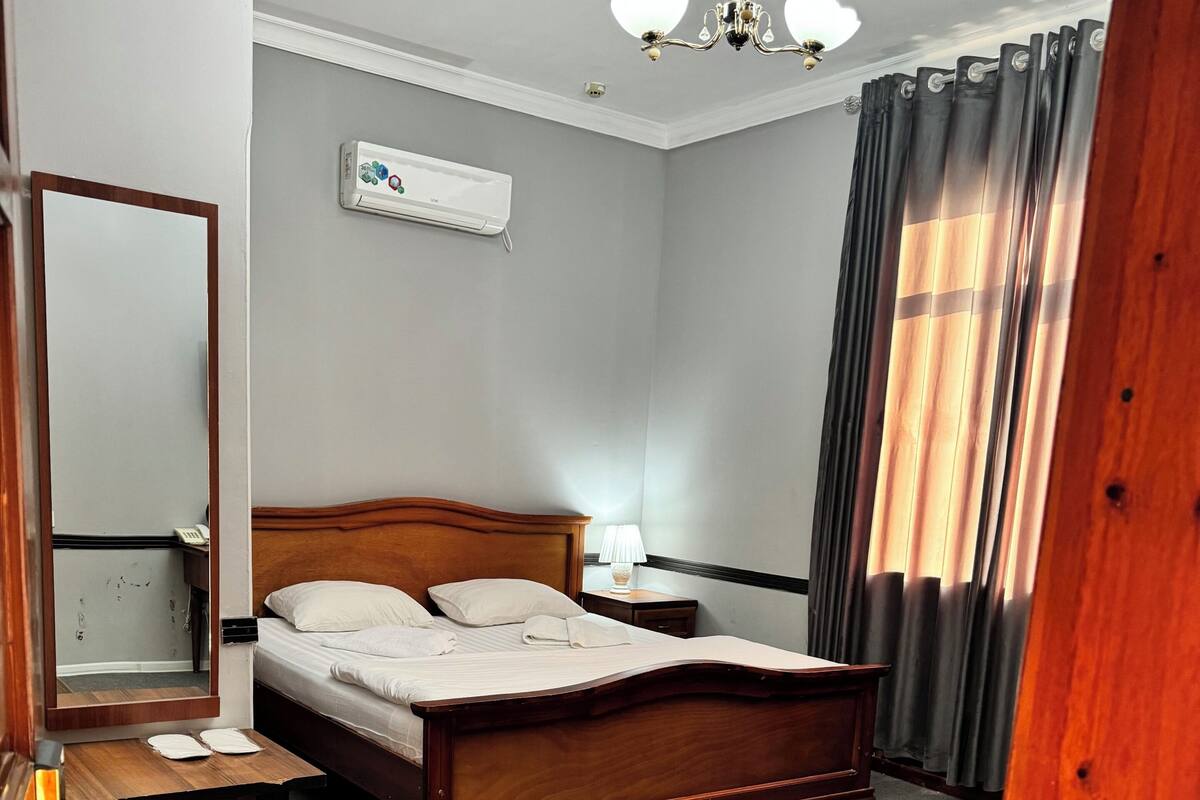 Quarto com cama de madeira, mesa de cabeceira, luminária, aparelho de ar condicionado e espelho.