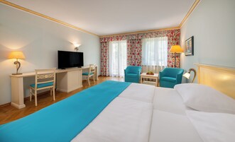 Quarto de hotel com cama, escrivaninha com televisão, duas cadeiras, mesa com frutas e cortinas florais.