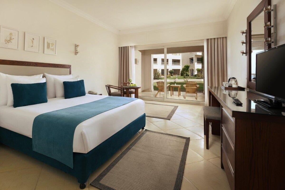 Quarto de hotel com cama grande, televisão, secretária e varanda com mobiliário exterior.