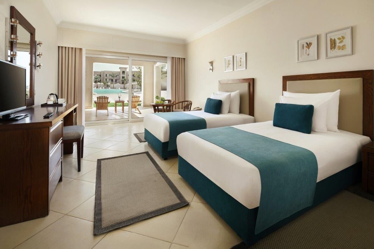 Quarto de hotel com duas camas, uma mesa, uma televisão e vista para uma piscina.