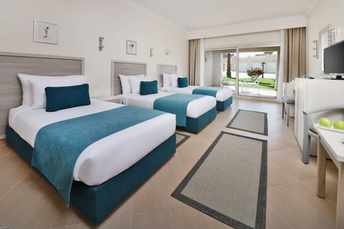 Quarto de hotel com três camas, televisão, escrivaninha e vista para o exterior.