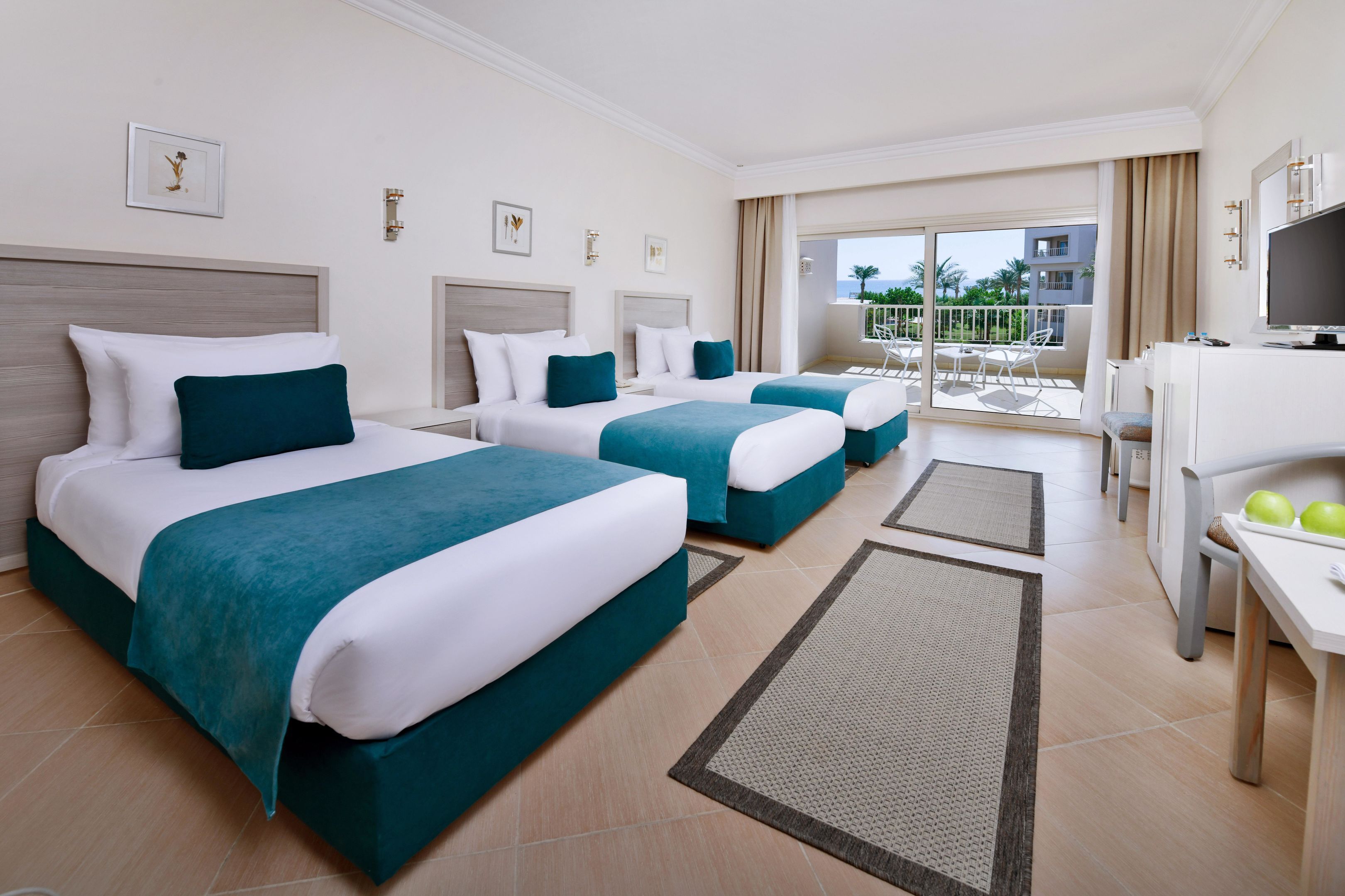 Quarto de hotel com duas camas, televisão, secretária com maçãs e varanda com vista.