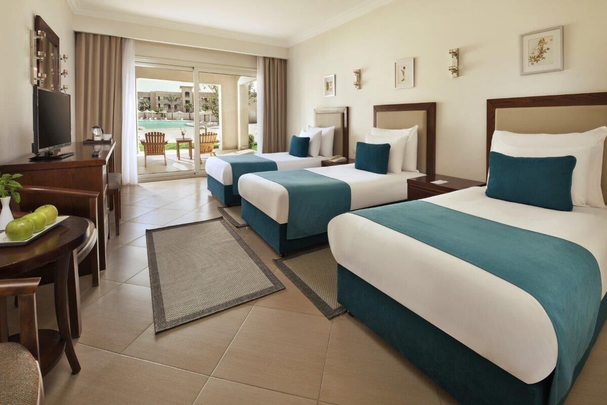 Quarto de hotel com duas camas, uma escrivaninha com televisão, área de refeições e vista para a piscina.