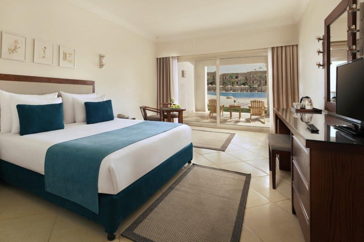Quarto de hotel com uma cama grande, uma escrivaninha, uma televisão e vista para uma piscina.