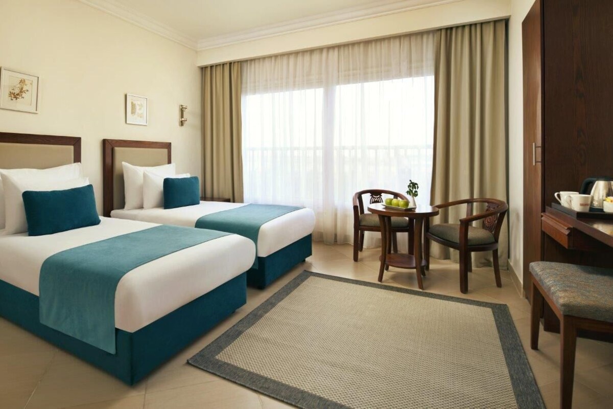Quarto de hotel com duas camas, uma escrivaninha com cadeira, uma mesinha com uma planta e uma janela com cortinas.
