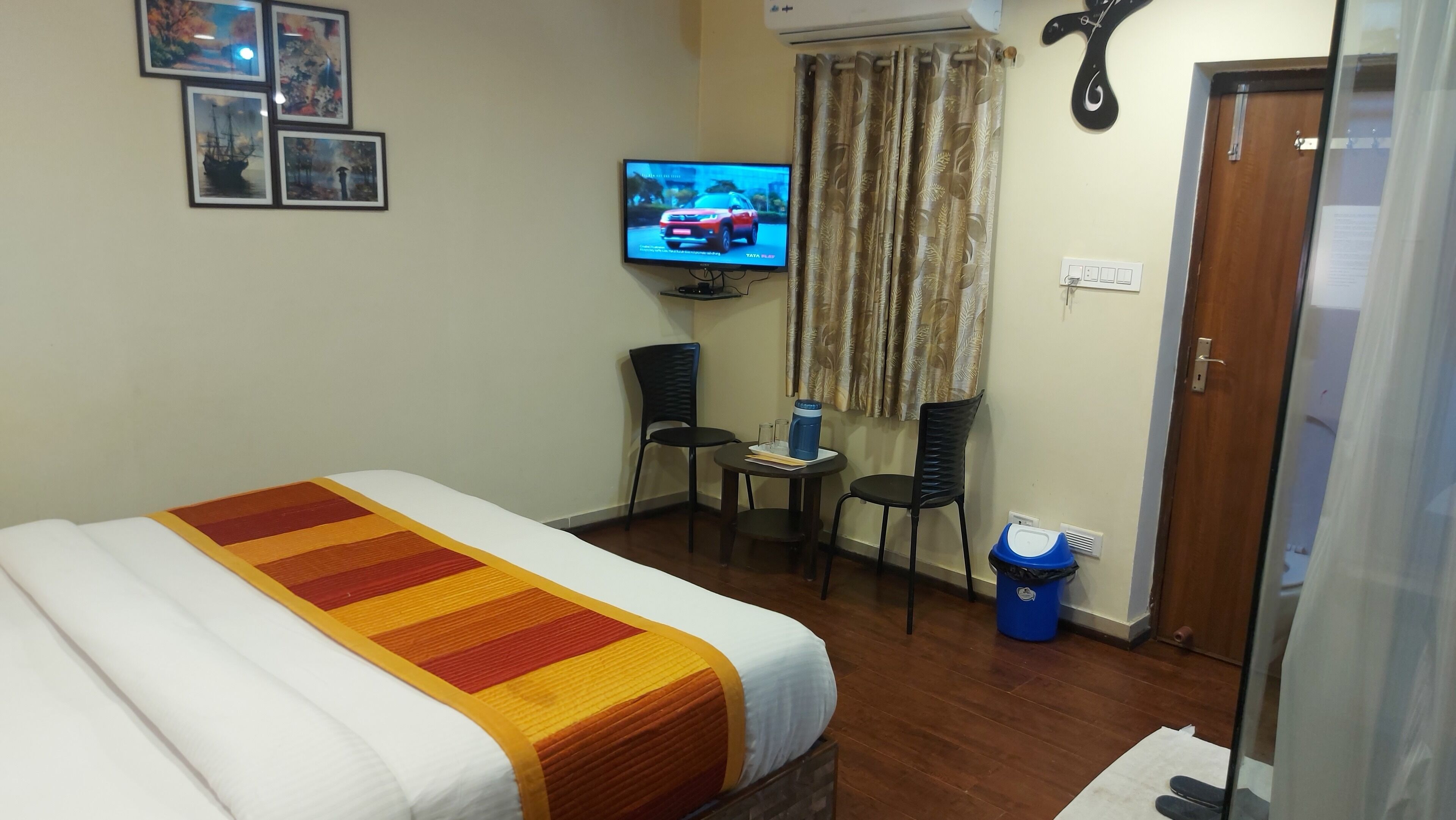 Quarto de hotel com uma cama, televisão, uma mesinha com cadeiras, janela com cortinas e porta.