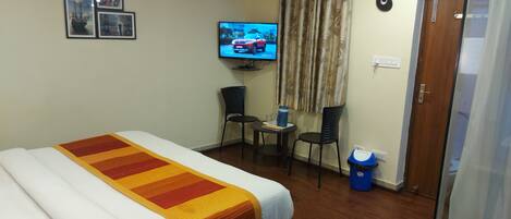 Quarto de hotel com uma cama, televisão, uma mesinha com cadeiras, janela com cortinas e porta.