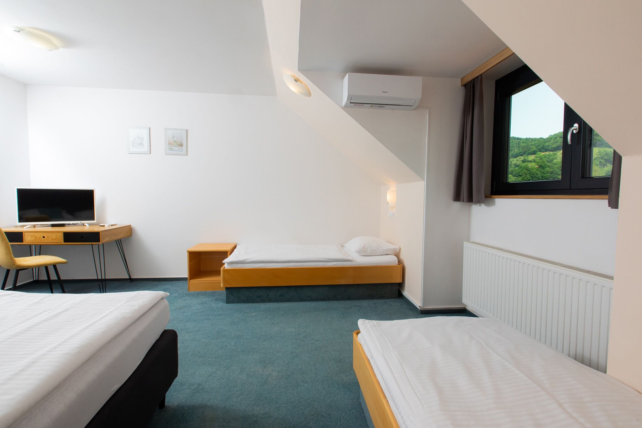 Quarto de hotel com duas camas, uma escrivaninha, uma cadeira, ar condicionado e uma janela com vista para o verde.