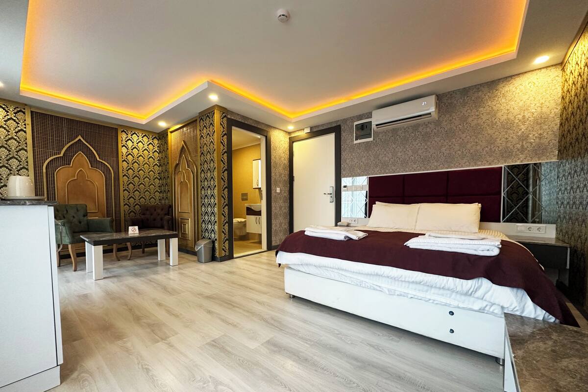Quarto de hotel moderno com uma cama grande, área de estar e banheiro visível por uma porta aberta.