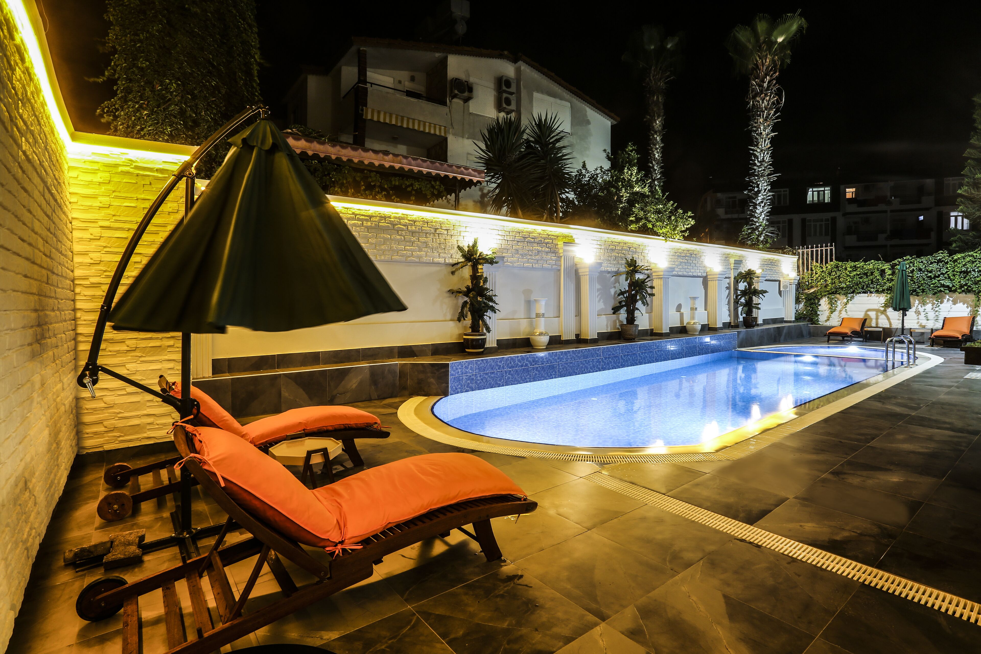Piscina com espreguiçadeiras e um abajur, cercada por um muro de tijolos e palmeiras.