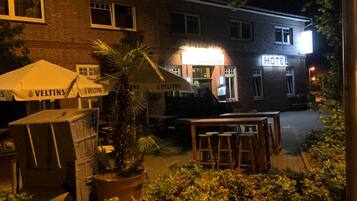 Exterior de um hotel à noite, com área de estar externa e guarda-sóis com a inscrição "VELTINS".