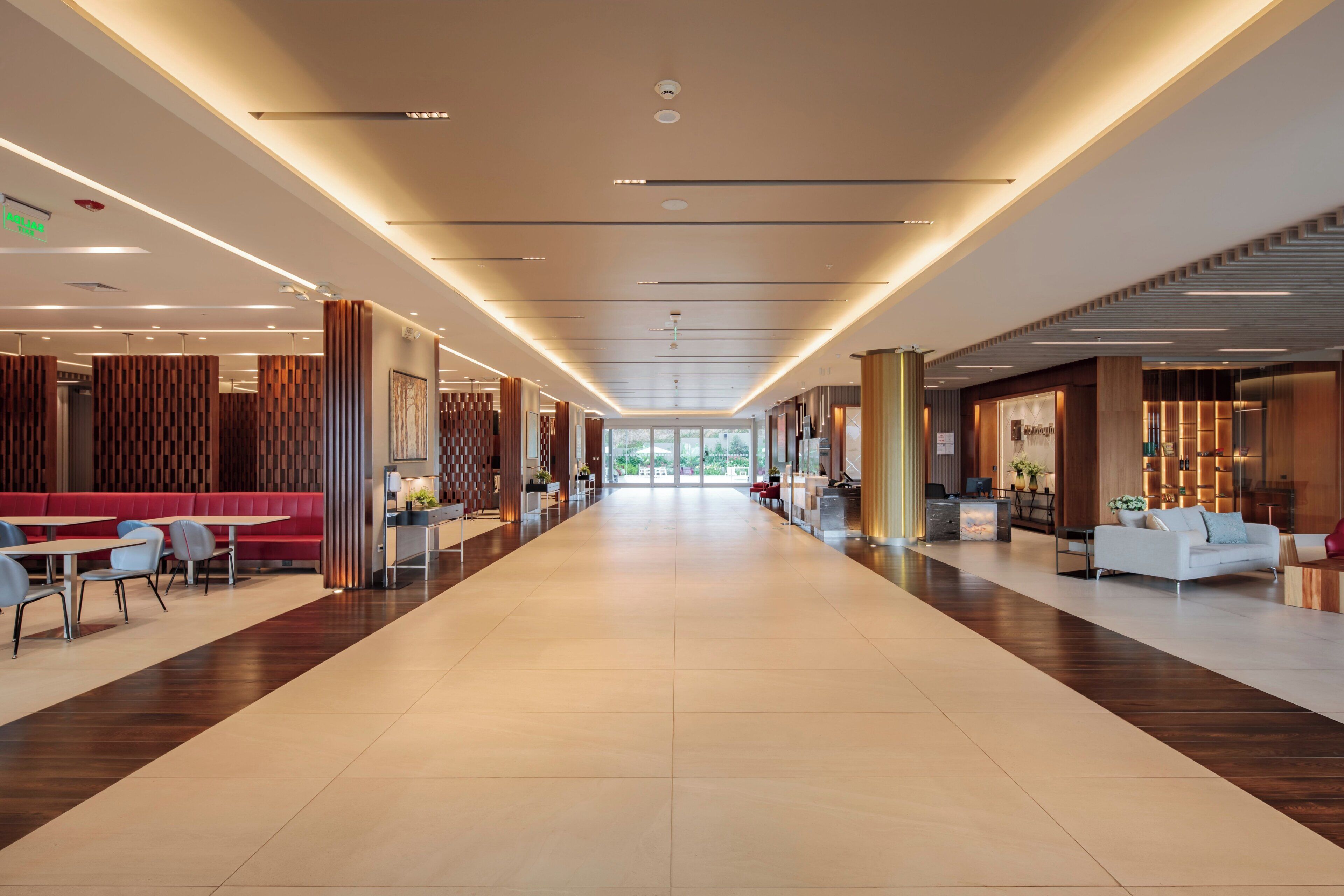 Corredor de hotel espaçoso com design moderno, apresentando um caminho central, áreas de estar em ambos os lados e uma receção no final.