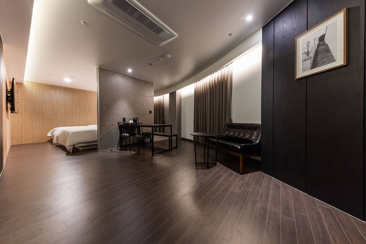 Quarto de hotel moderno com cama, escrivaninha, cadeira, sofá e uma grande janela com cortinas.