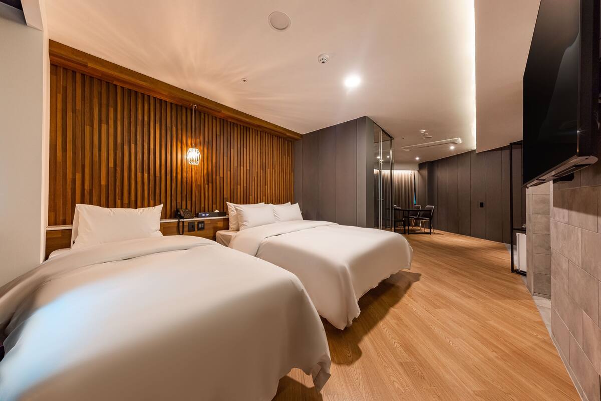 Quarto de hotel moderno com duas camas, painel de parede de madeira, televisão de tela plana e uma pequena mesa com cadeiras.