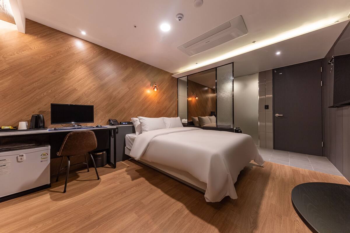 Quarto de hotel moderno com piso de madeira, cama com roupa de cama branca, escrivaninha com computador e um pequeno frigorífico.