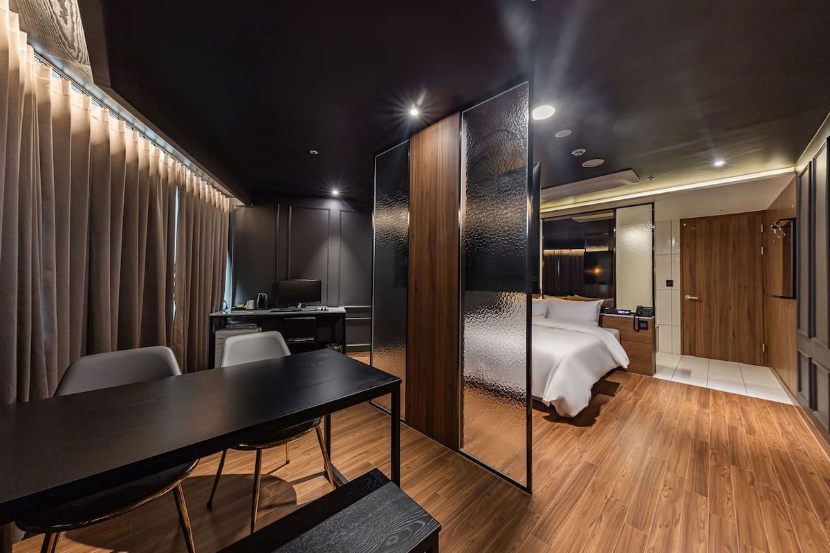 Quarto de hotel moderno com piso de madeira, uma escrivaninha com computador, uma cama e um espelho grande.