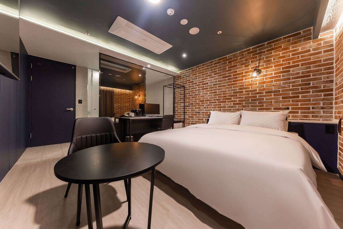 Quarto de hotel moderno com parede de tijolos, uma cama grande, uma escrivaninha com cadeira e uma pequena mesa redonda.