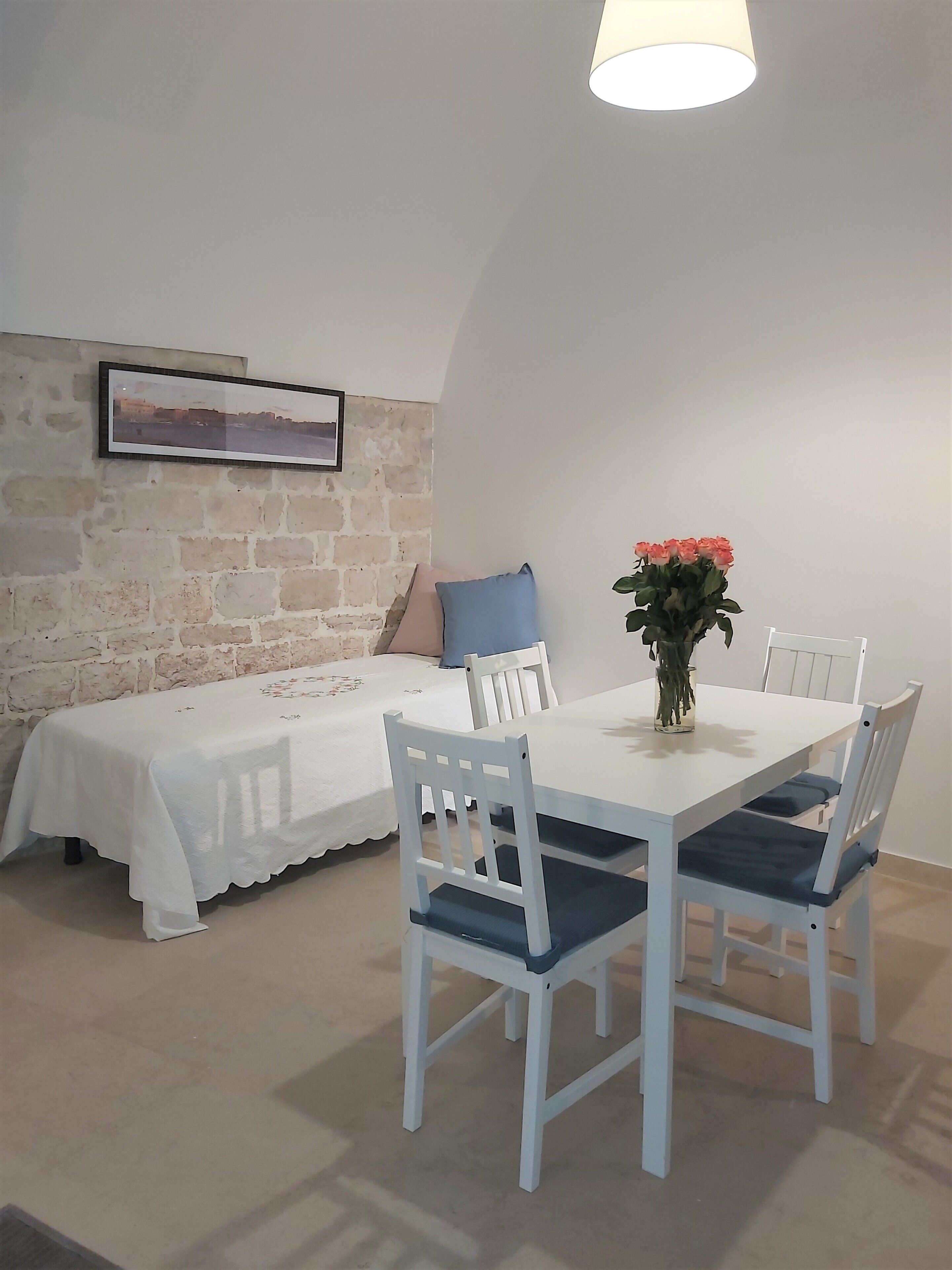 Quarto com mesa e cadeiras brancas, uma cama com cobertura branca, travesseiro azul, quadro emoldurado na parede e um vaso com flores sobre a mesa.