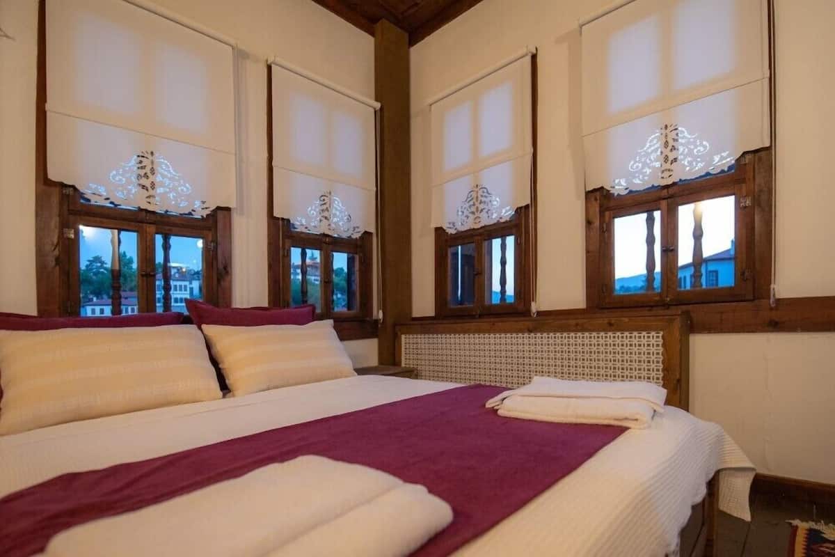 Quarto com uma cama grande, cabeceira de madeira e janelas com cortinas decorativas.