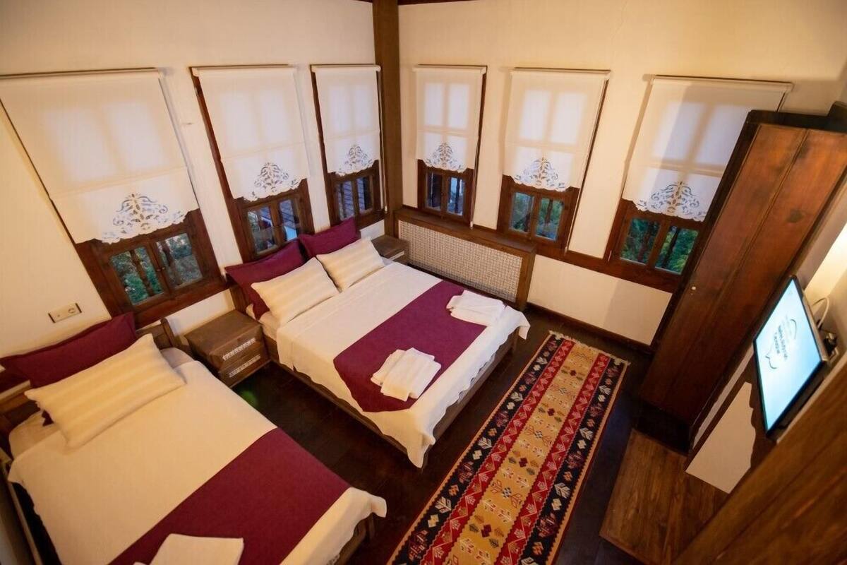 Quarto de hotel com quatro camas, cada uma com colcha vermelha e branca, cabeceira de madeira e janela com cortina de renda.