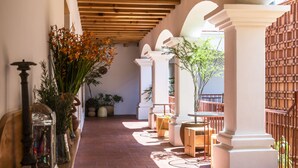 Uma área externa coberta com bancos e mesas de madeira, plantas em vasos e piso de azulejos.
