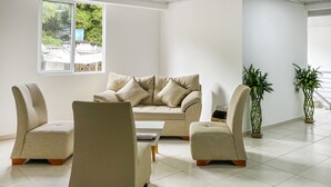 Uma sala de estar moderna com um sofá bege, duas poltronas e uma mesa de centro. Há duas plantas em vasos e uma janela com vista para o verde lá fora.