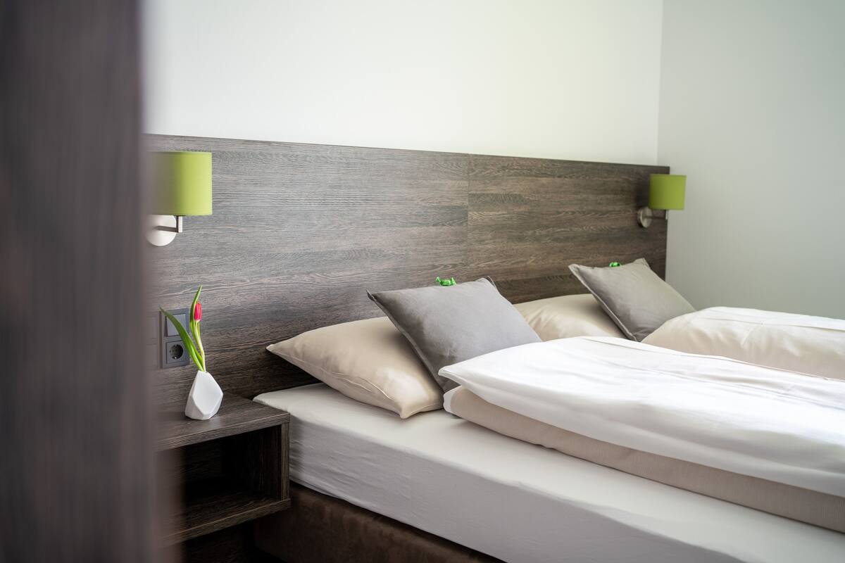 Cama bem arrumada com duas almofadas e um vaso com uma flor, cabeceira de madeira e duas luminárias fixadas na parede.