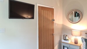 Quarto com porta de madeira, televisão de tela plana na parede, mesa de cabeceira com abajur e espelho sobre a mesa.
