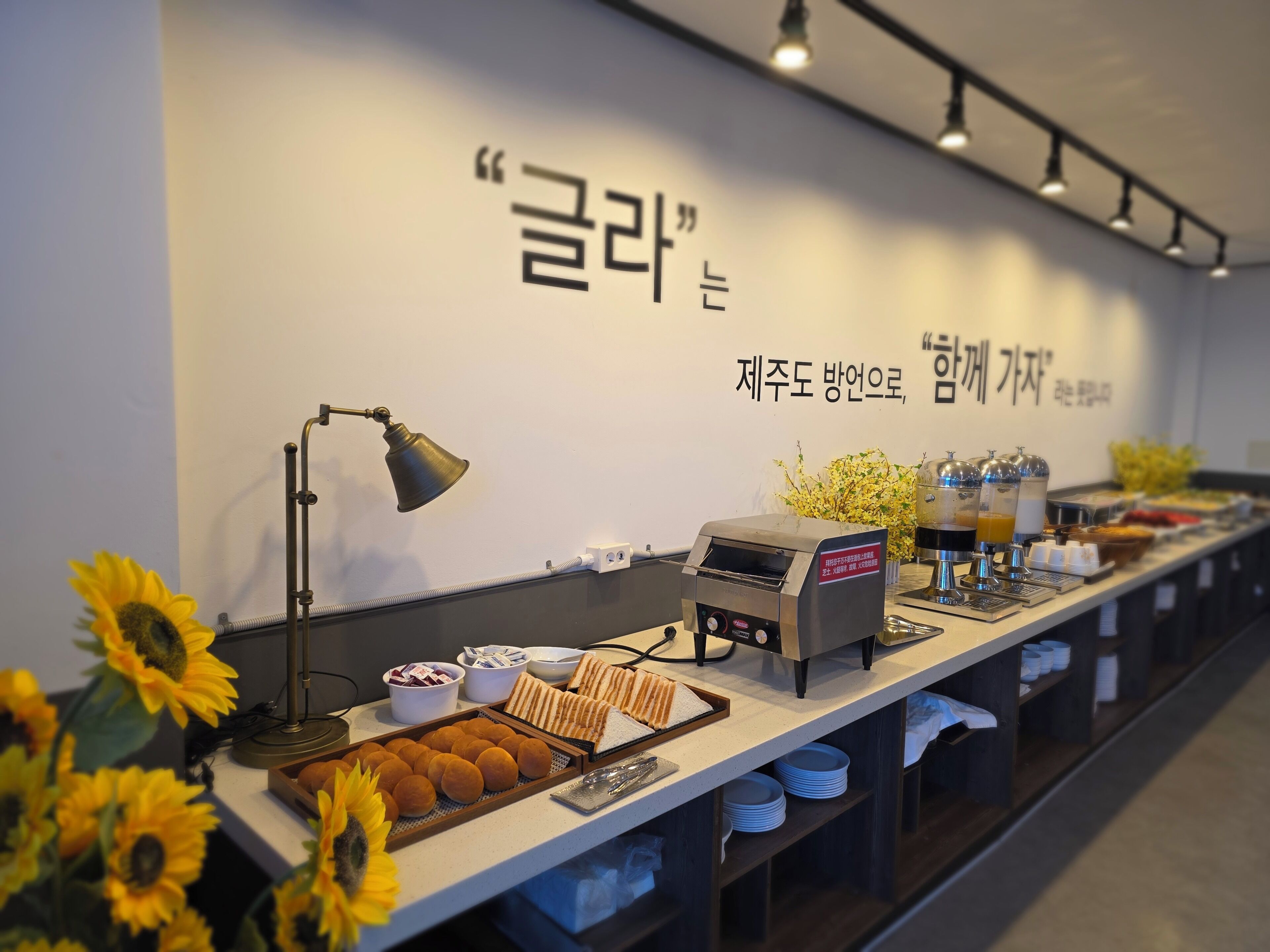 Um buffet de café da manhã com diversos alimentos, uma torradeira e uma parede com texto em coreano.