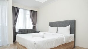 Quarto de hotel moderno com uma cama grande, cabeceira estofada cinza, roupa de cama branca e uma poltrona cinza.