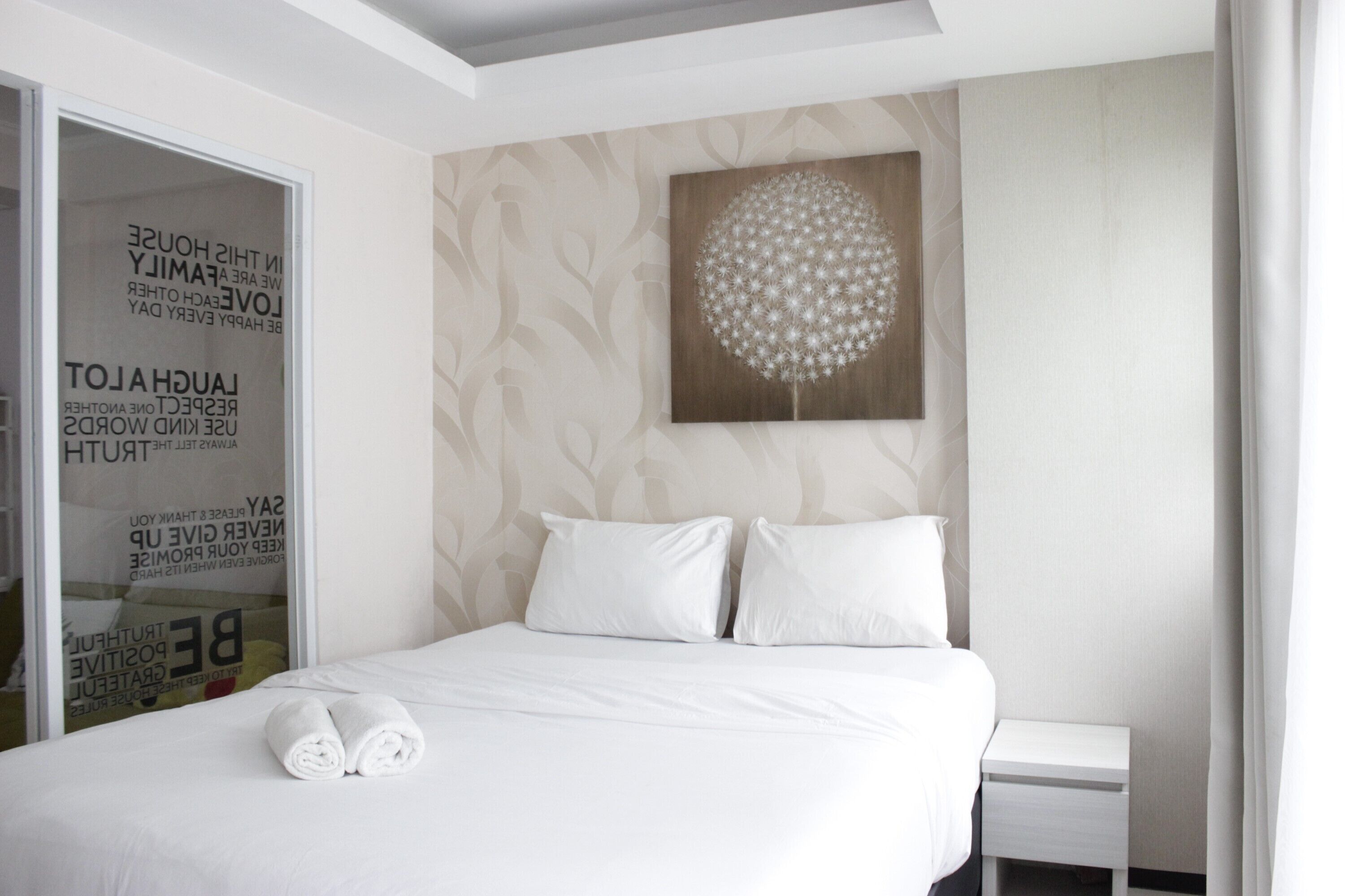 Cama bem arrumada com lençóis e almofadas brancos, uma obra de arte na parede e um espelho com texto motivacional.