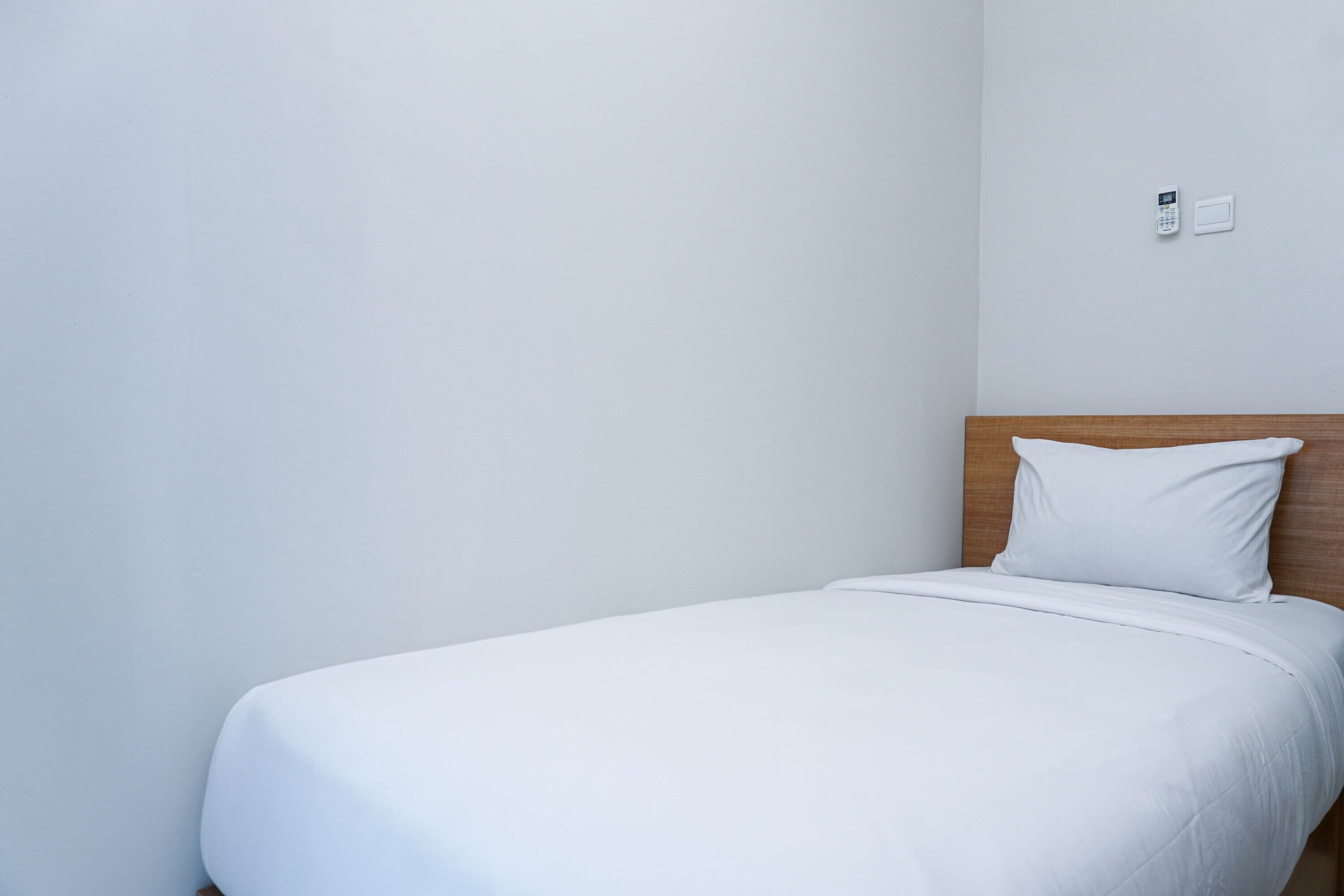 Uma cama de solteiro com roupa de cama branca em um quarto com paredes brancas lisas e uma cabeceira de madeira.