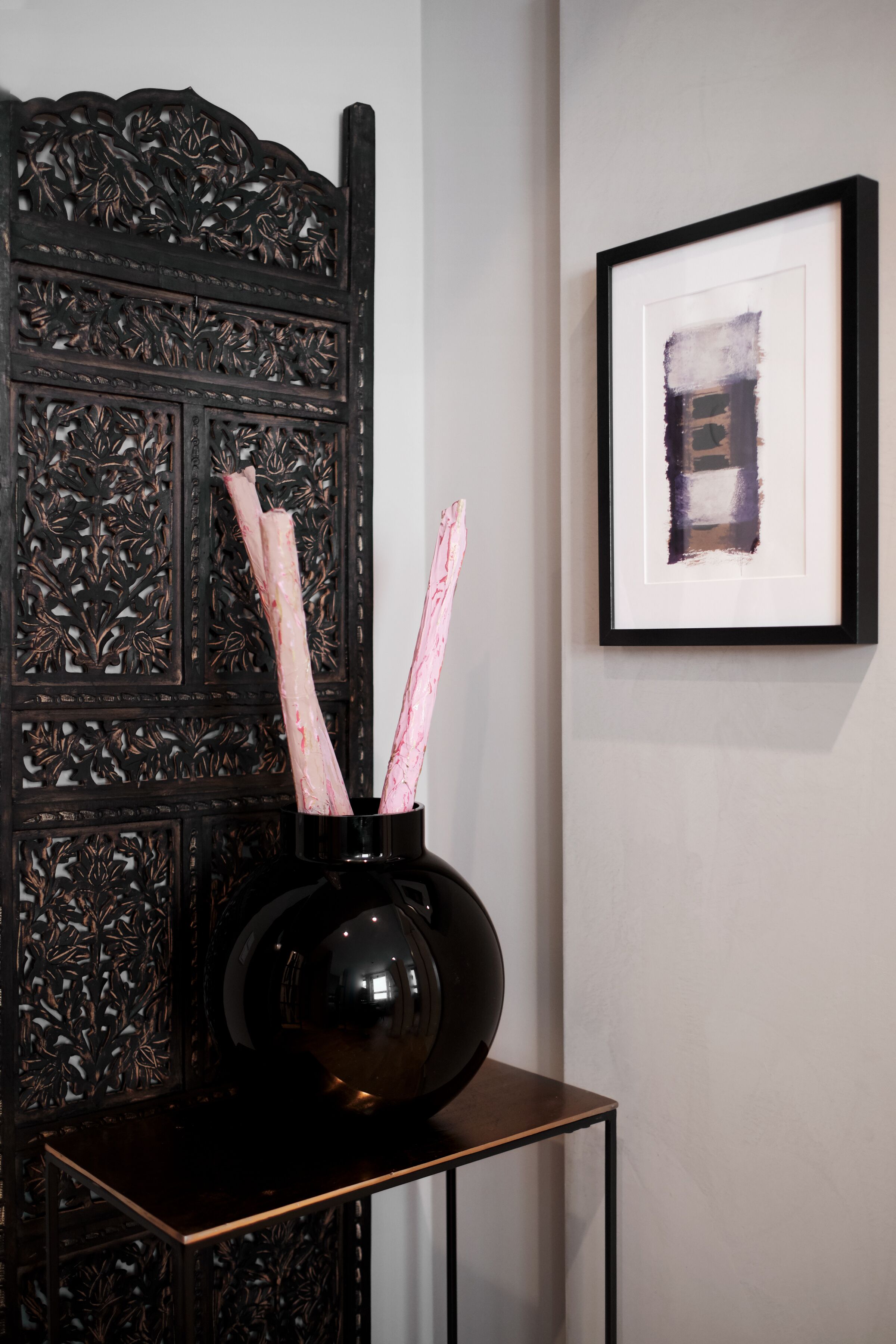 Um vaso preto com gravetos rosa e brancos listrados sobre uma mesa preta, em frente a uma parede com um painel ornamentado e uma pintura abstrata emoldurada.