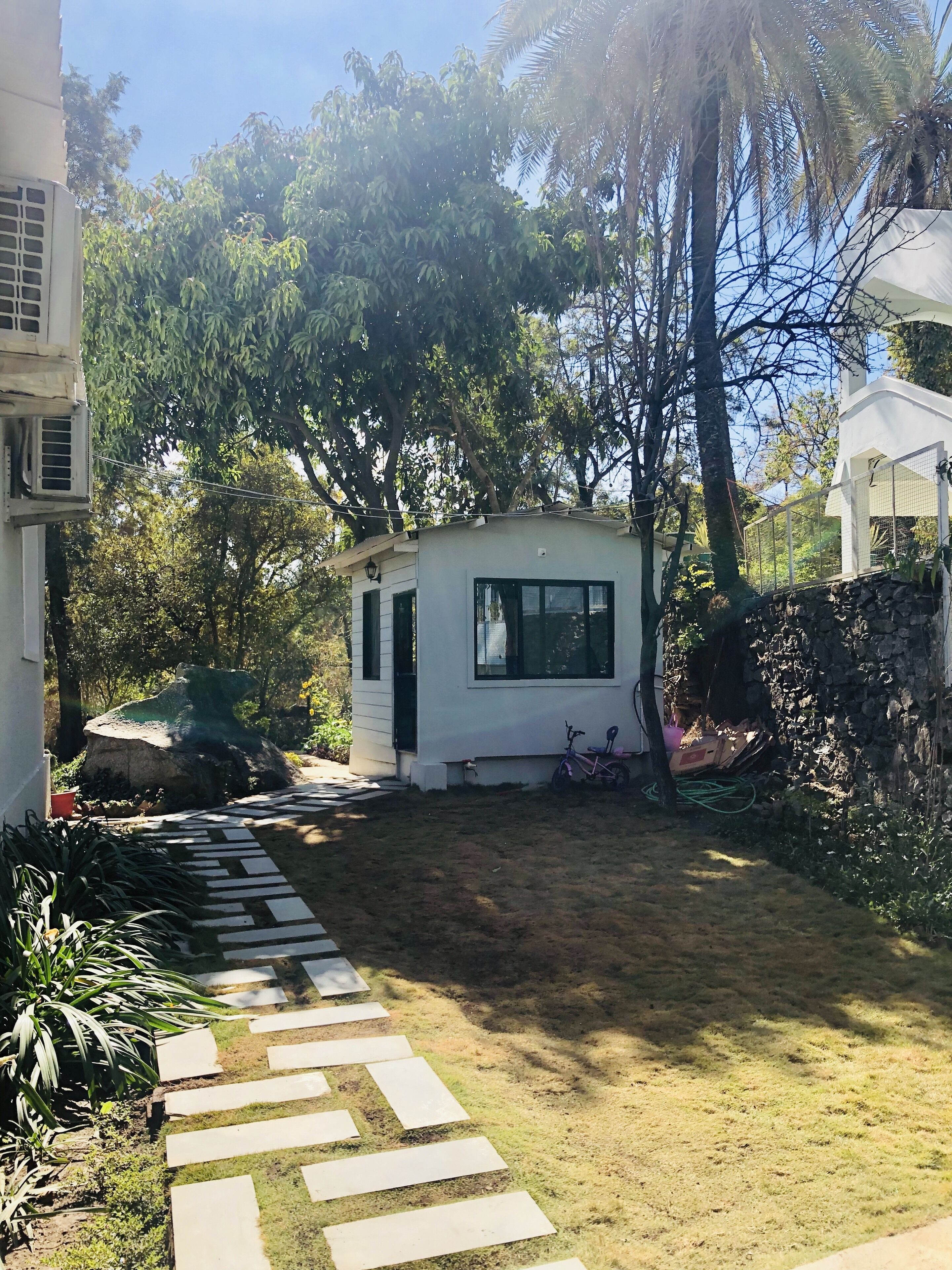 Uma pequena casa branca com um caminho de pedras que a leva, rodeada por árvores e vegetação.
