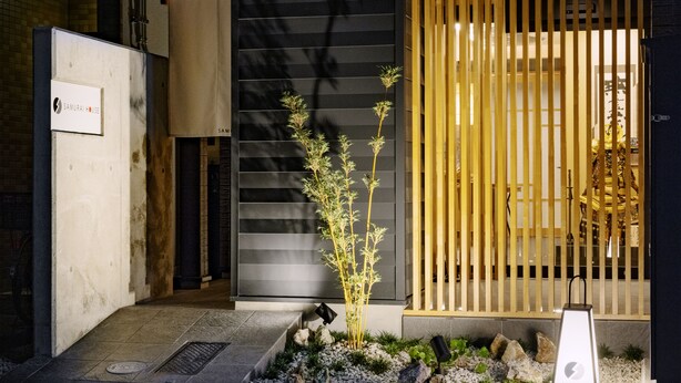 Uma fachada moderna e escura, porta de ripas de madeira e uma pequena área ajardinada com pedras e plantas.