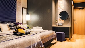 Quarto de hotel moderno com uma cama grande, uma mesa de cabeceira com abajur, um banquinho pequeno e um espelho fixado na parede.