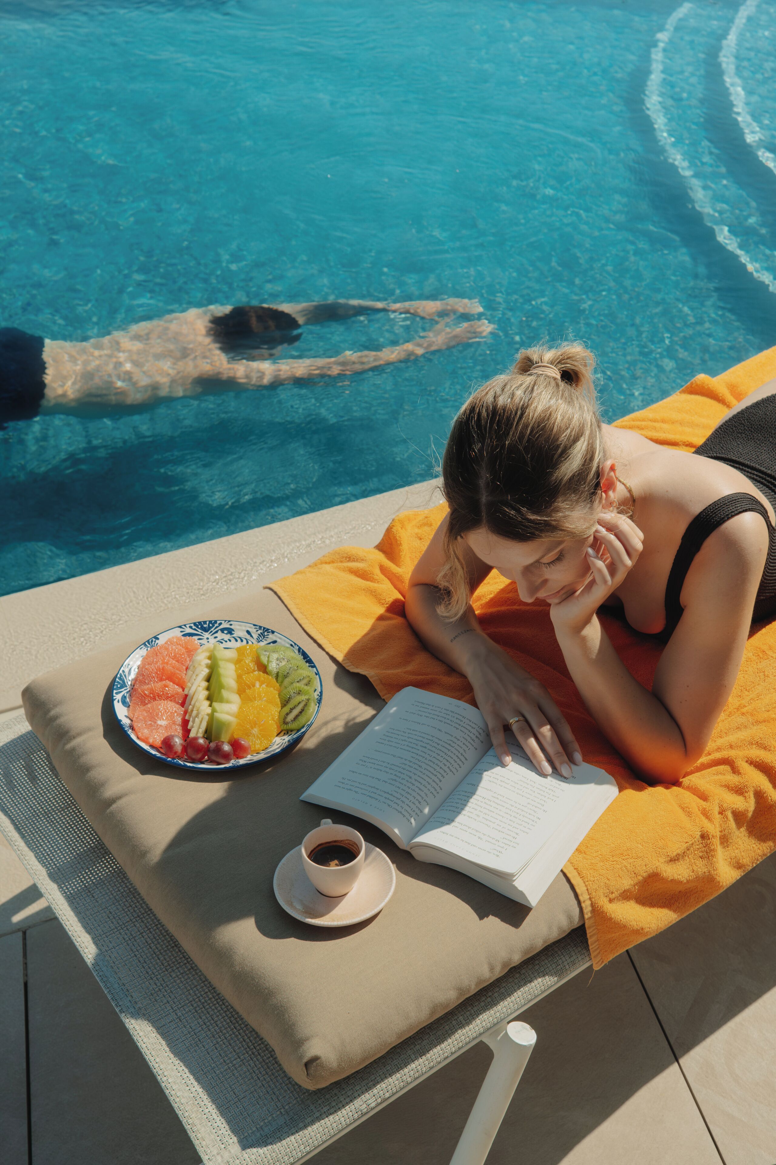 Uma pessoa relaxando em uma espreguiçadeira à beira da piscina, com um livro, uma tábua de frutas e uma xícara de café.