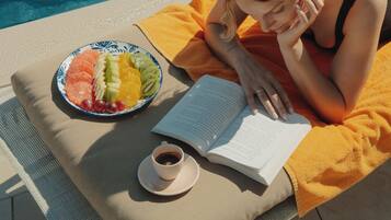 Uma pessoa relaxando em uma espreguiçadeira à beira da piscina, com um livro, uma tábua de frutas e uma xícara de café.