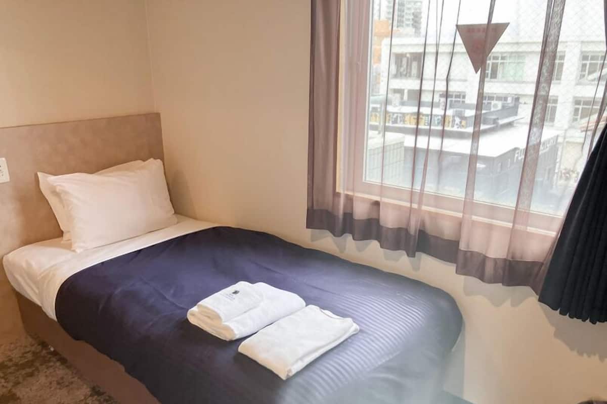 Uma cama de solteiro com colcha azul escura e almofadas brancas, uma janela com cortinas finas e vista para prédios lá fora.